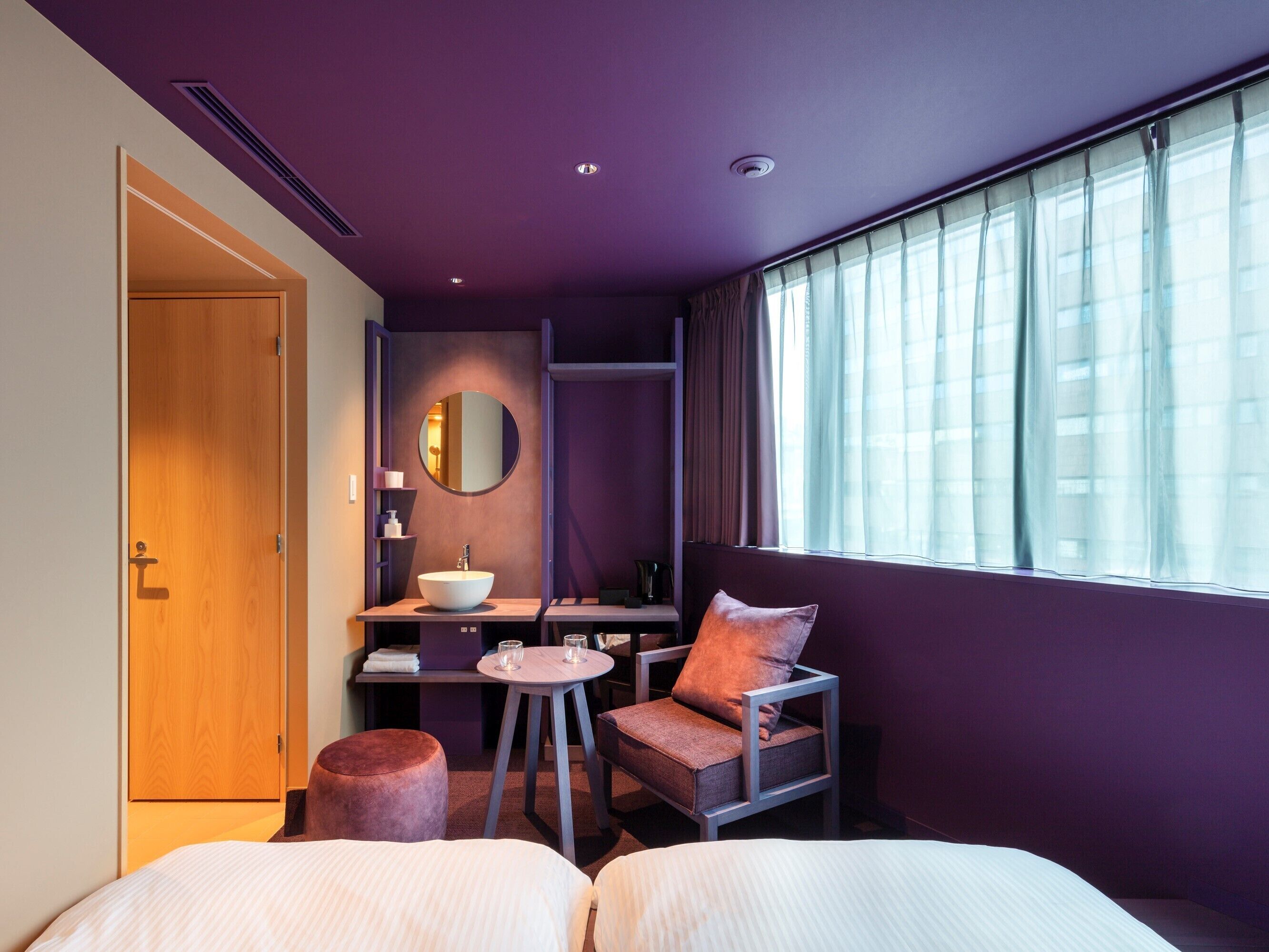 Quarto de hotel moderno com uma cama grande, uma mesa redonda pequena, uma cadeira, um sofá, um banheiro com pia e espelho e uma janela com cortinas.