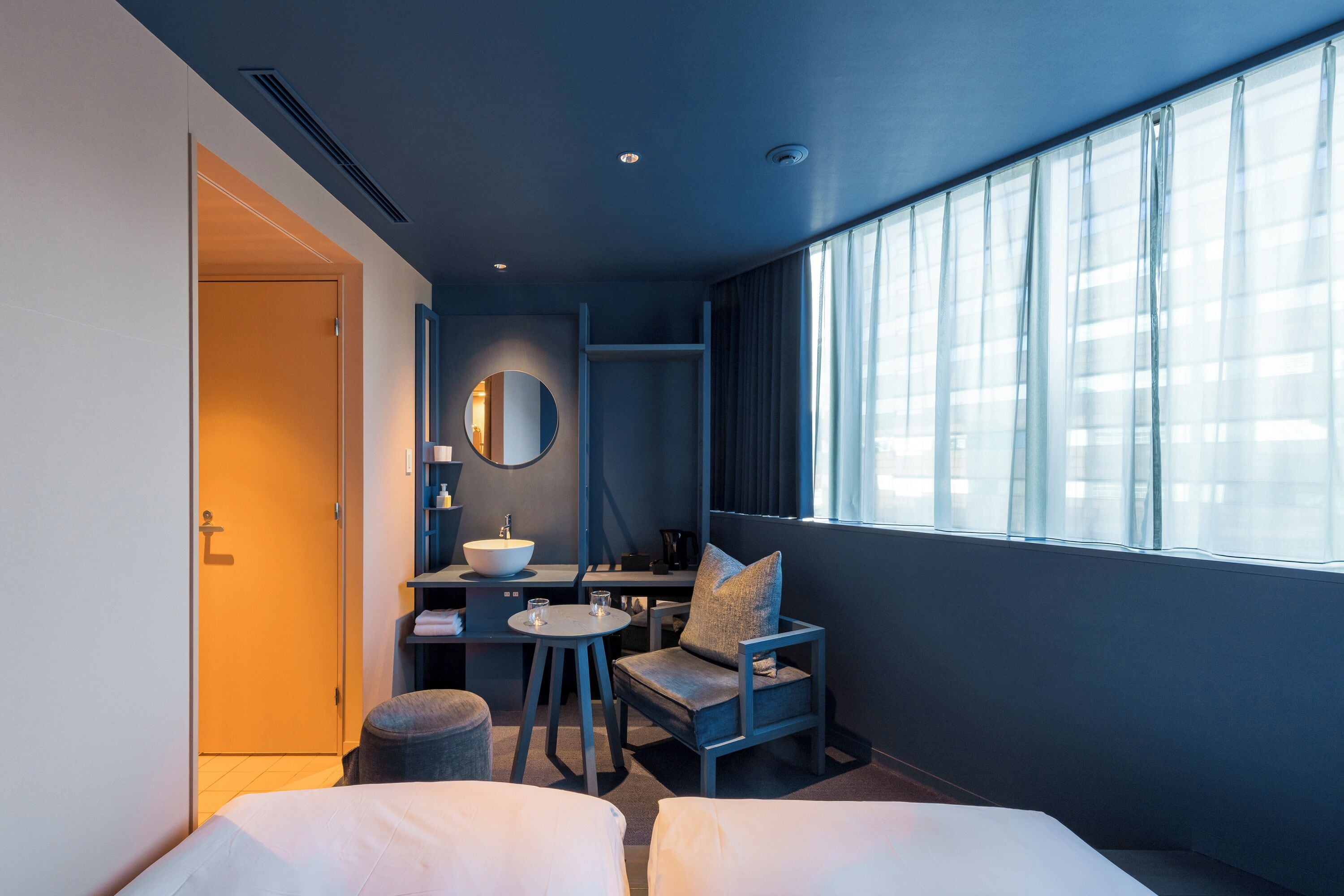 Quarto de hotel moderno com uma cama, uma pequena escrivaninha com cadeira, um espelho redondo e uma janela com cortinas.