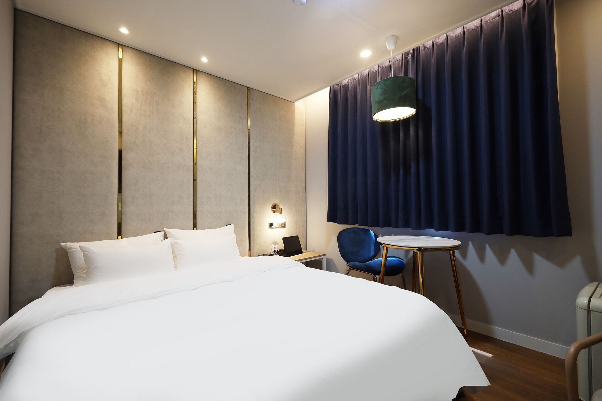 Quarto de hotel com uma cama grande, uma escrivaninha com um laptop, uma cadeira, uma mesinha e uma janela com cortinas.