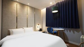Quarto de hotel com uma cama grande, uma escrivaninha com um laptop, uma cadeira, uma mesinha e uma janela com cortinas.
