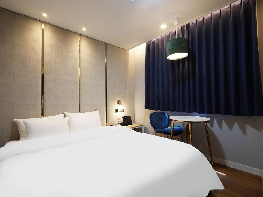 Quarto de hotel com uma cama grande, uma escrivaninha com um laptop, uma cadeira, uma mesinha e uma janela com cortinas.