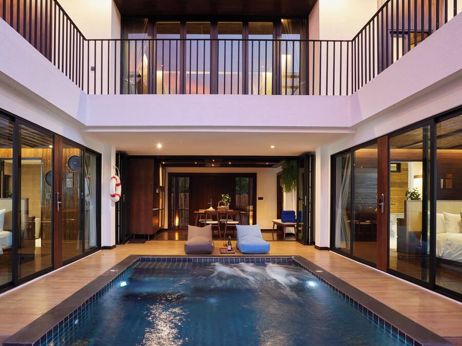 Uma área interna de piscina moderna com barreira de vidro, piso de madeira e um espaço de estar com cadeiras e uma mesa.