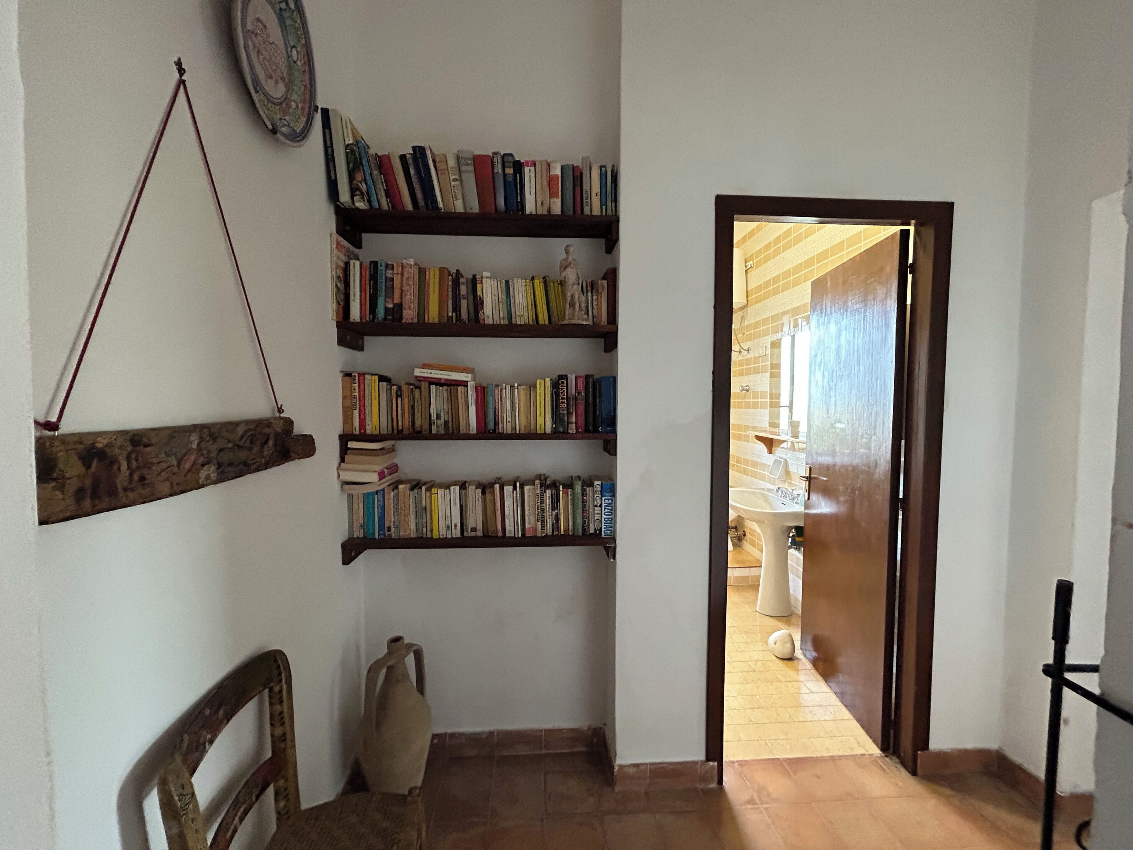 Um corredor estreito com uma estante cheia de livros, uma cadeira rústica e uma porta que dá para um banheiro.