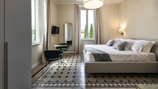 Quarto de hotel moderno com uma cama grande, uma escrivaninha e uma cadeira. Há uma televisão fixada na parede e uma janela com cortinas.