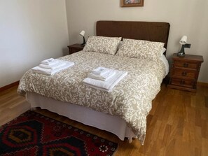 Uma cama bem arrumada com colcha estampada e almofadas, uma mesinha de cabeceira com abajur e um criado-mudo de madeira.
