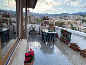 Uma varanda com mesa e cadeiras, plantas em vasos e vista para uma paisagem urbana com montanhas ao fundo.