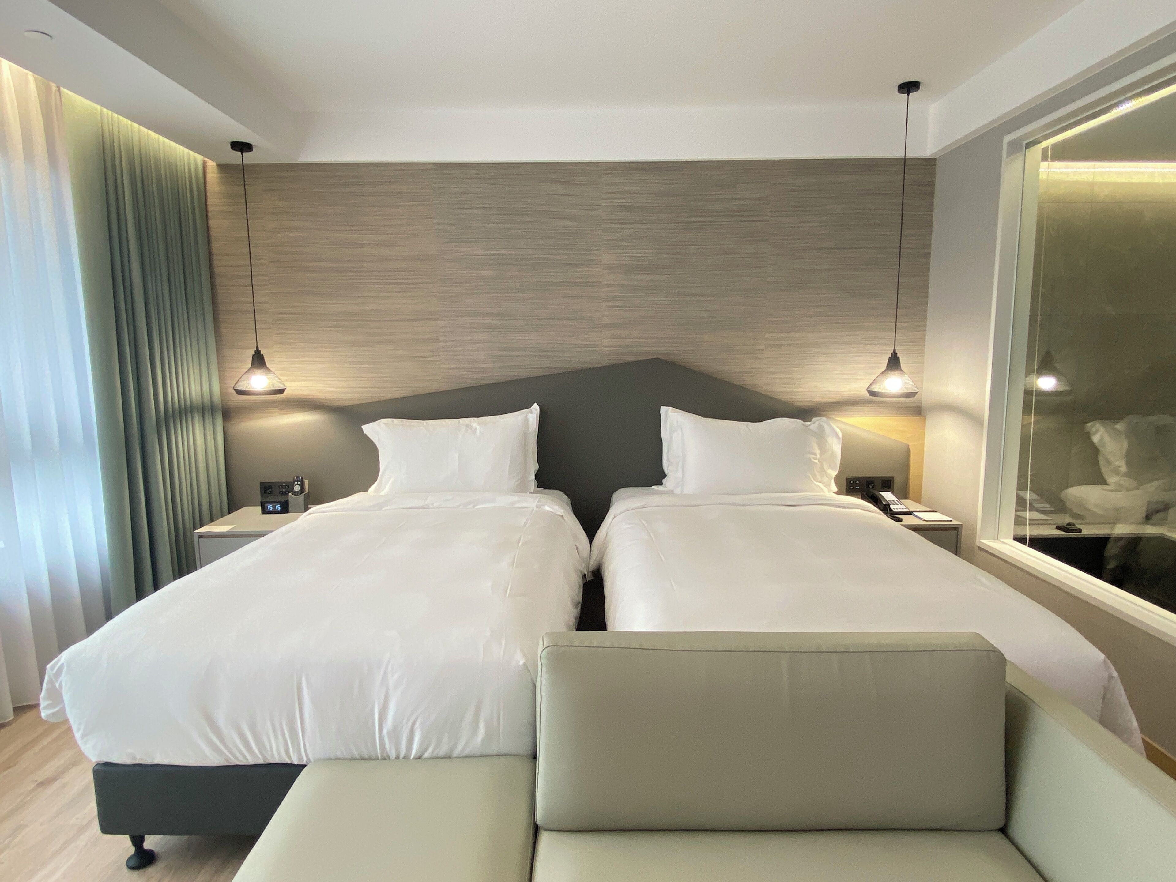 Quarto de hotel com duas camas, um sofá, uma mesinha de cabeceira com telefone e uma janela grande.