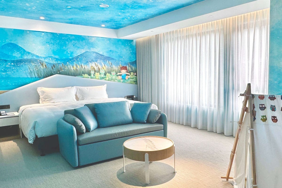 Quarto de hotel moderno com uma cama grande, um sofá azul, uma mesa de centro redonda e um mural de uma criança à beira-mar.