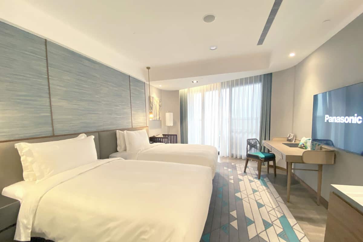 Quarto de hotel com duas camas, uma escrivaninha, uma cadeira, televisão e uma janela grande com cortinas.