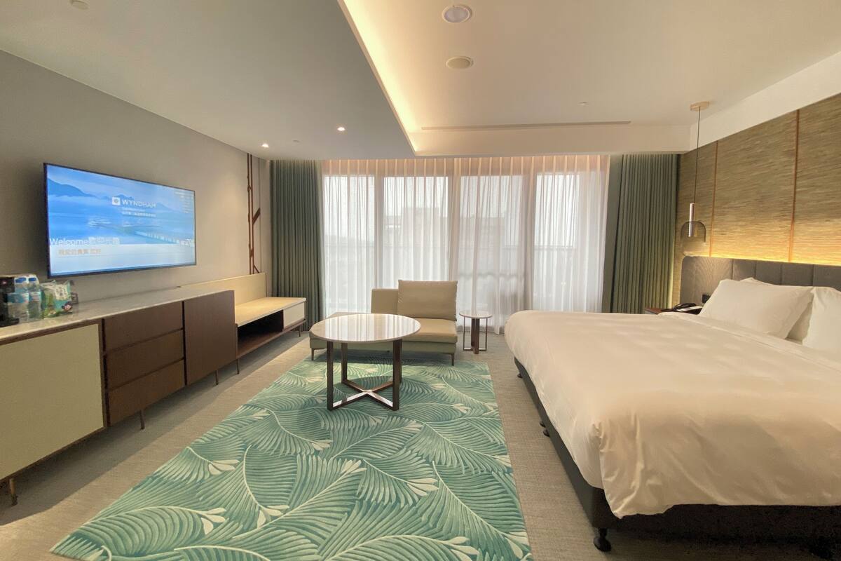 Quarto de hotel com cama, sofá, televisão na parede e escrivaninha com computador.