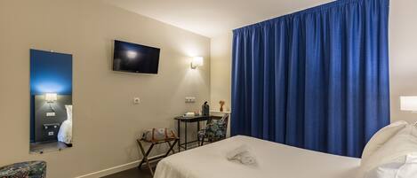 Quarto de hotel com cama, escrivaninha, cadeira, televisão e uma grande janela com cortinas.