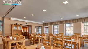 Uma ampla sala de jantar com várias mesas e cadeiras de madeira, um aparador de madeira com portas de vidro e grandes janelas que permitem a entrada de luz natural.