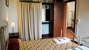Um quarto com cama, mesa de cabeceira, luminária, janela com cortinas, espelho e porta para outro cômodo.