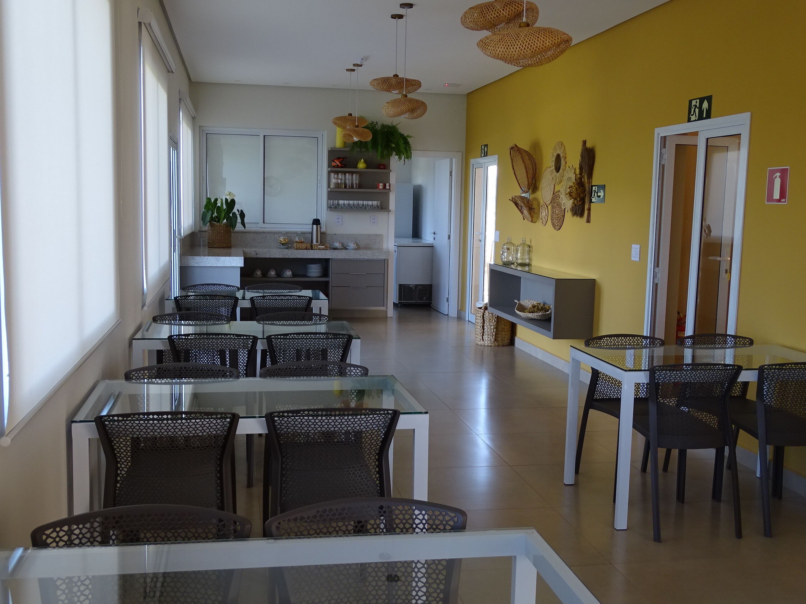 Uma sala de jantar ampla com móveis modernos, uma cozinha ao fundo e uma parede de destaque amarelo vibrante.