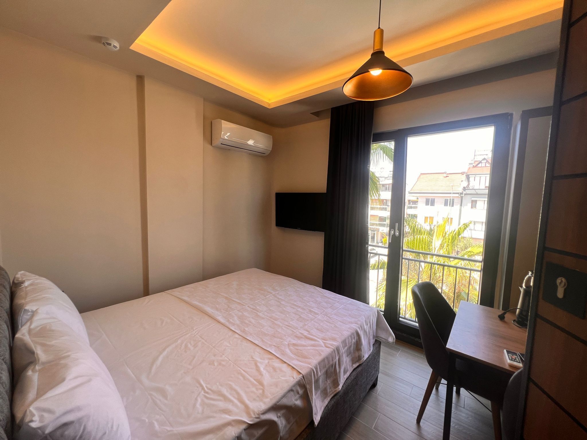 Quarto de hotel com cama, escrivaninha, cadeira, aparelho de ar condicionado, televisão e varanda com vista para edifícios e árvores.
