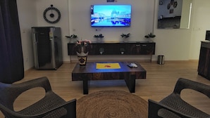 Uma sala de estar moderna com uma TV de tela plana na parede, uma mesa de centro de madeira escura, duas cadeiras pretas, um tapete tecido e uma pequena geladeira.