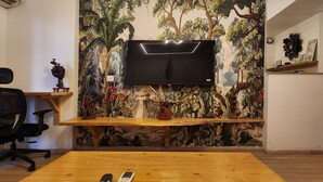 Uma sala de estar com televisão de tela plana na parede, exibindo um mural tropical, uma mesa de centro de madeira com controles remotos e uma cadeira de escritório preta.