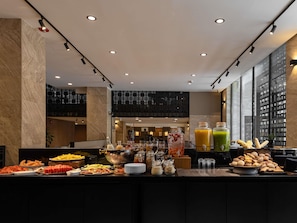 Buffet com diversos alimentos e bebidas, interior moderno com teto alto e iluminação embutida, e vista para a cidade através de grandes janelas.