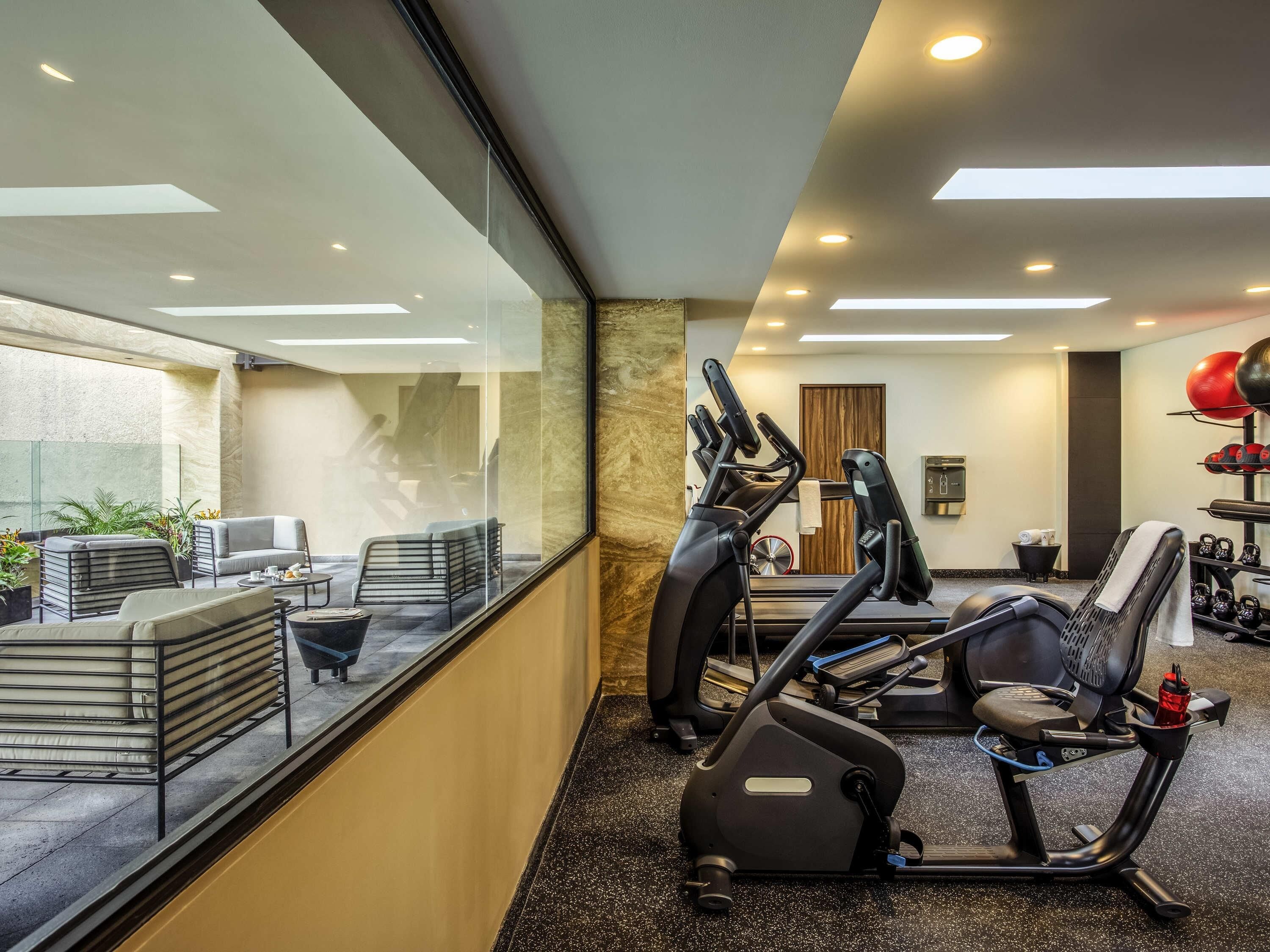 Uma academia moderna com equipamentos de ginástica, área de estar com cadeiras e uma mesa, e vista através de um painel de vidro.