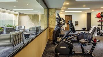 Uma academia moderna com equipamentos de ginástica, área de estar com cadeiras e uma mesa, e vista através de um painel de vidro.