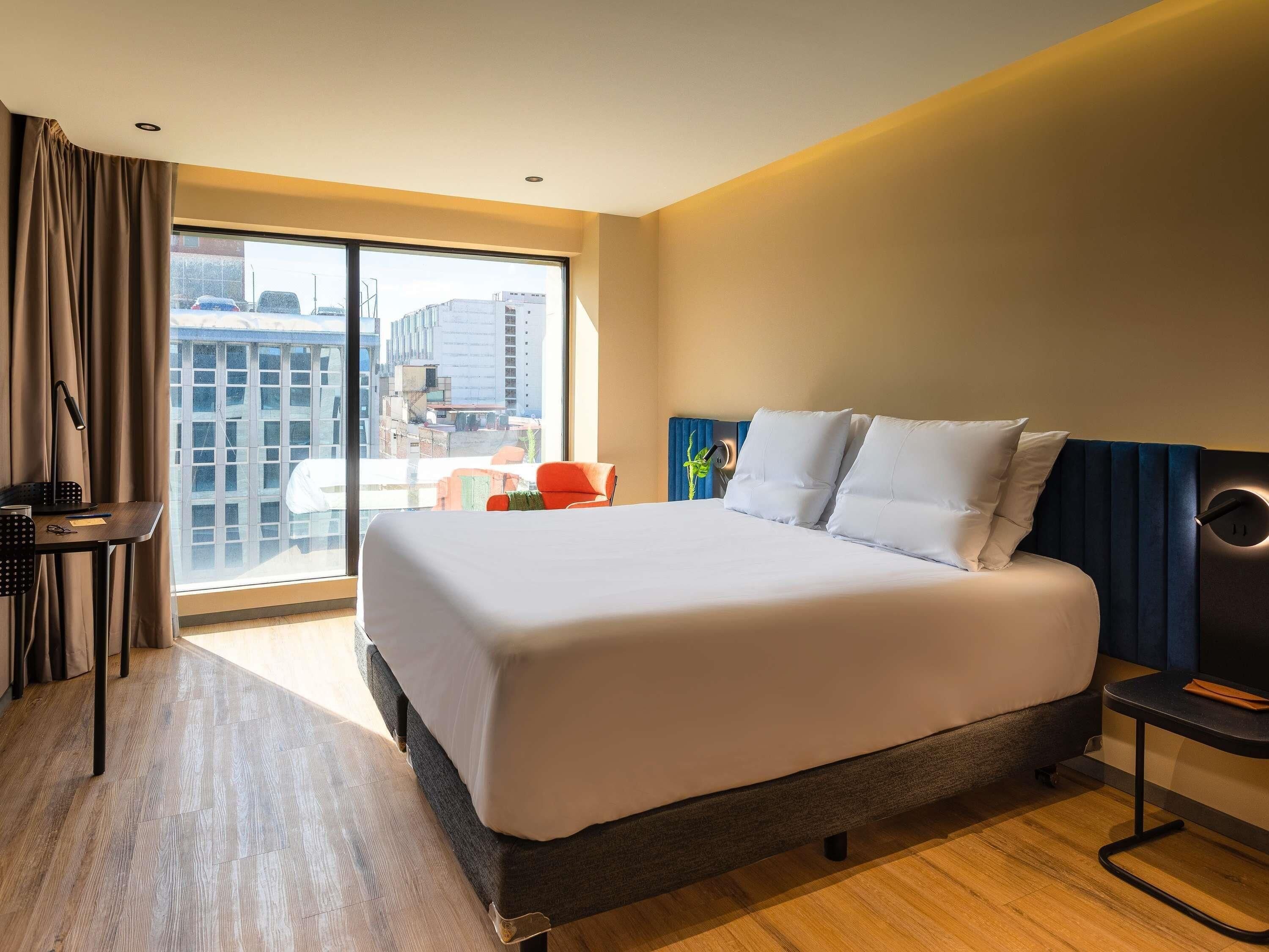 Quarto de hotel com uma cama grande, uma escrivaninha, uma cadeira e vista da cidade através de uma janela ampla.