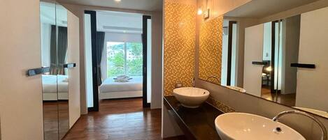 Casa de banho moderna com duas pias, um espelho grande e vista para um quarto com cama e cortinas.