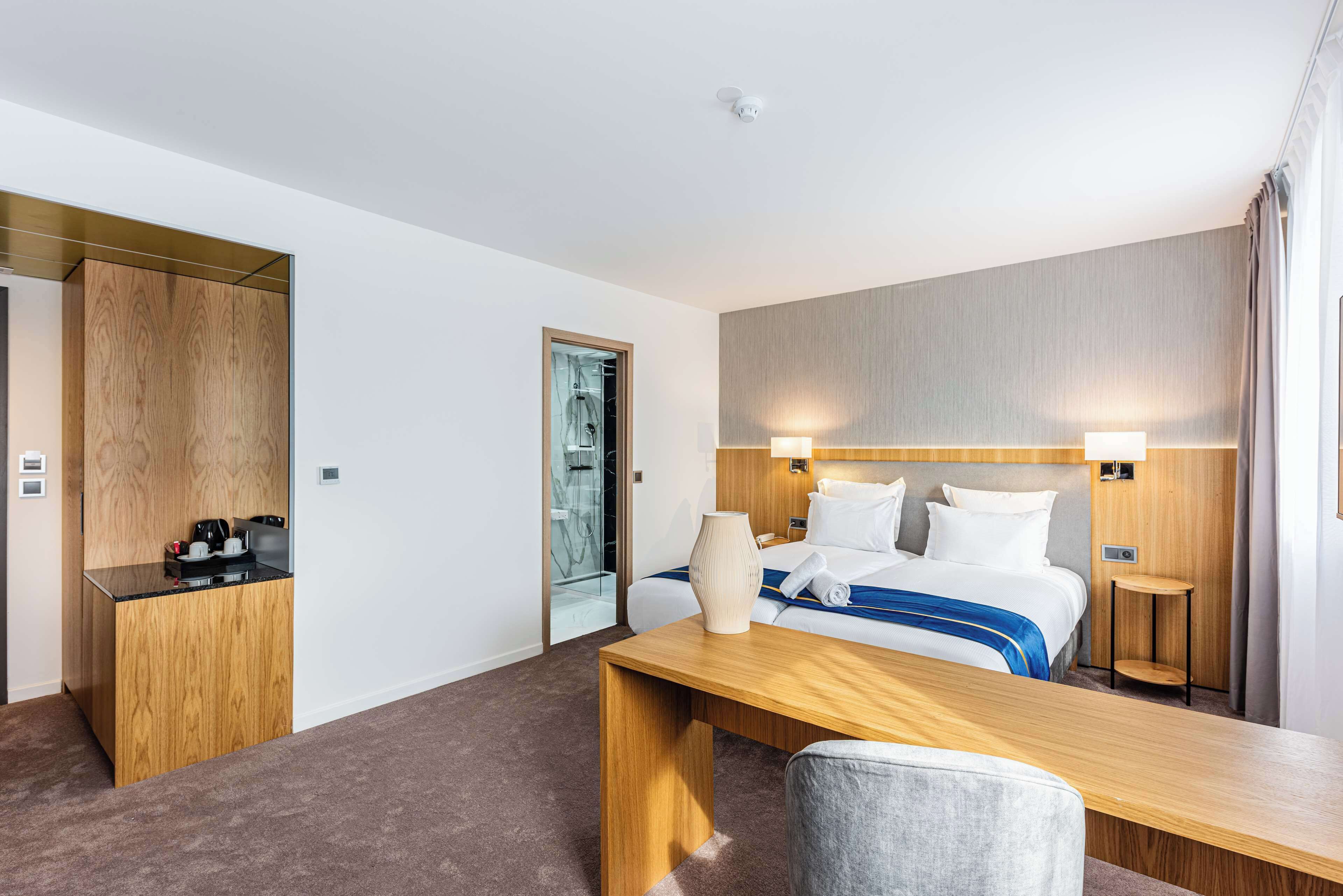 Quarto de hotel com cama, mesas de cabeceira, escrivaninha, cadeira, guarda-roupa e banheiro visível por uma porta de vidro.