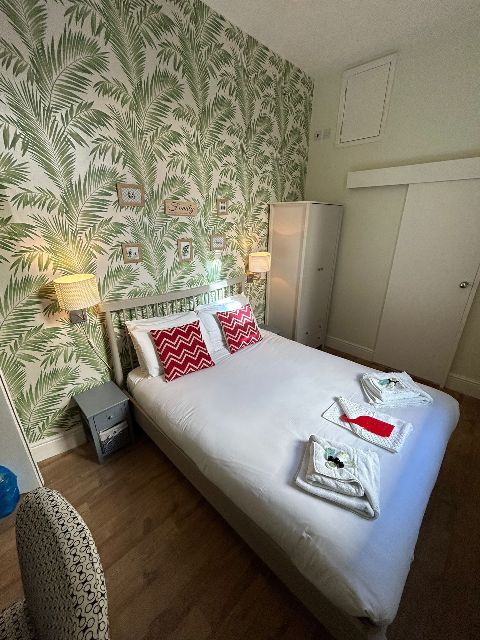 Cama bem arrumada com lençóis brancos e almofadas vermelhas, mesa de cabeceira com abajur, guarda-roupa e parede com padrão de folhagem tropical.
