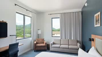 Quarto de hotel com cama, sofá, escrivaninha com cadeira, televisão e amplas janelas com vista para áreas verdes e edifícios.