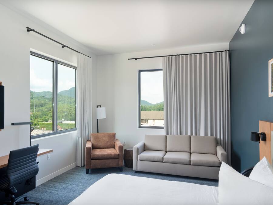 Quarto de hotel com cama, sofá, escrivaninha com cadeira, televisão e amplas janelas com vista para áreas verdes e edifícios.