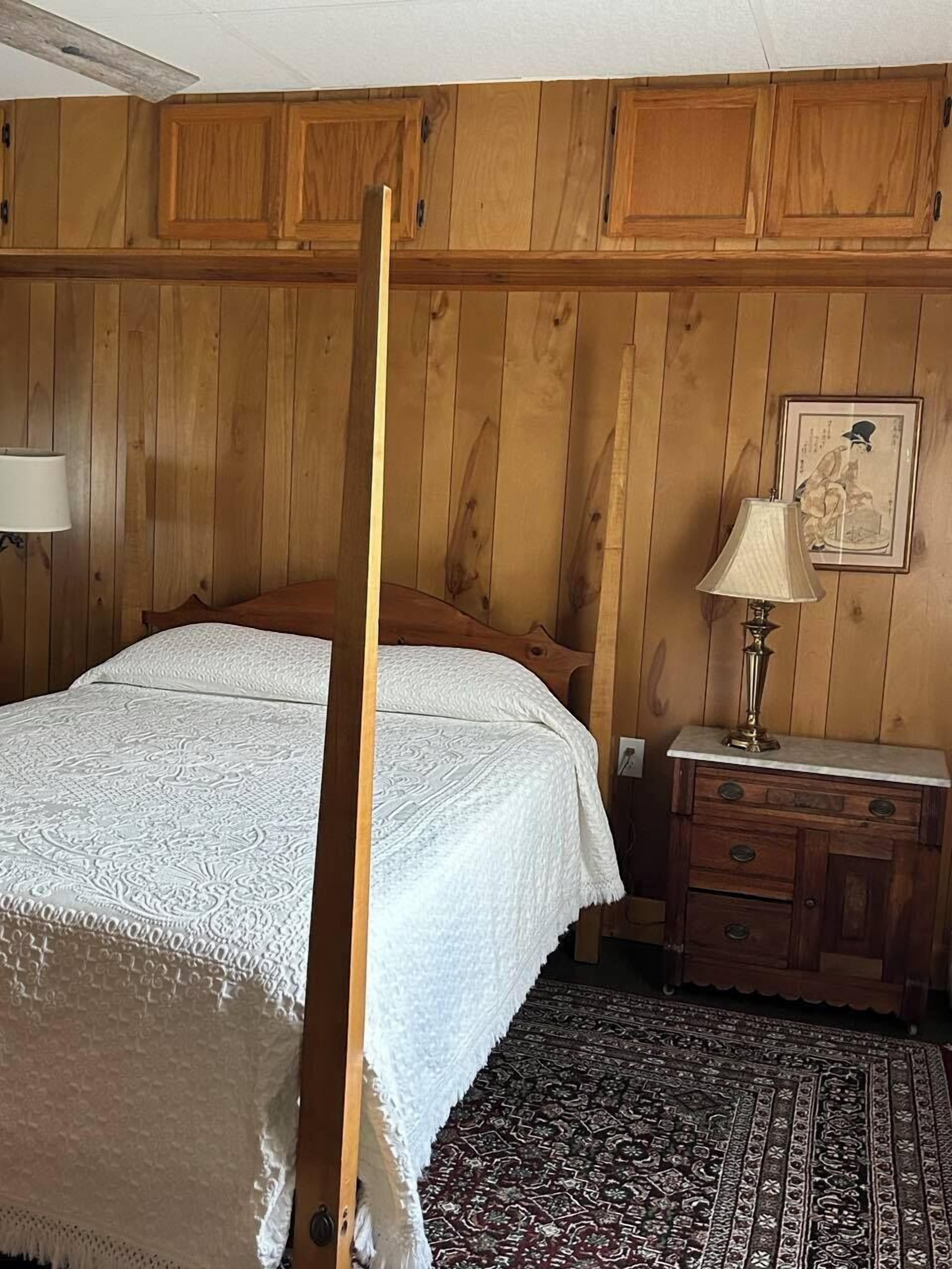 Quarto com cama de dossel, painéis de madeira nas paredes, mesa de cabeceira com abajur e tapete estampado.