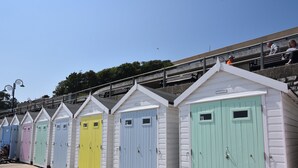 Uma fileira de cabanas de praia coloridas com portas numeradas, uma escadaria que leva a uma plataforma de observação e um céu claro.