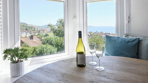 Um quarto com uma mesa de madeira, uma garrafa de vinho e duas taças. Um vaso com planta está no parapeito da janela, e uma almofada azul está no assento da janela.