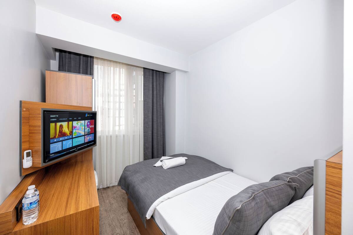 Quarto de hotel com televisão de tela plana, escrivaninha de madeira, garrafa de água, cama com roupa de cama cinza e janela com cortinas.