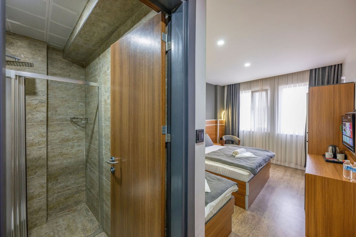 Quarto de hotel com chuveiro, cama, escrivaninha com televisão e janela com cortinas.