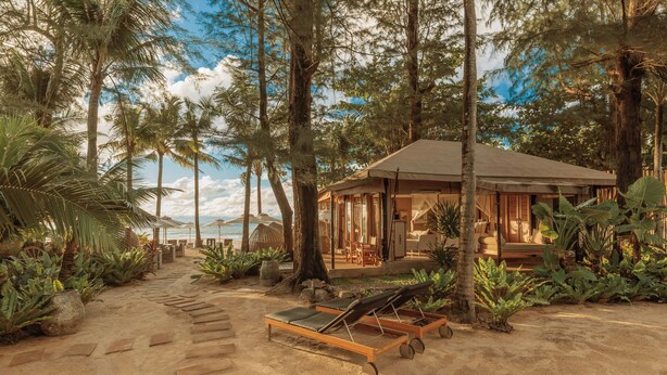 Um resort à beira-mar com um deque de madeira que leva a uma área de estar com espreguiçadeiras, cercado por vegetação tropical e uma grande árvore.