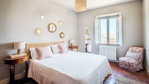 Quarto com cama, mesas de cabeceira, uma cadeira, janela e decorações nas paredes.