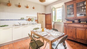 Cozinha com armários brancos, uma mesa de madeira posta para refeições e vista para o exterior pela janela.