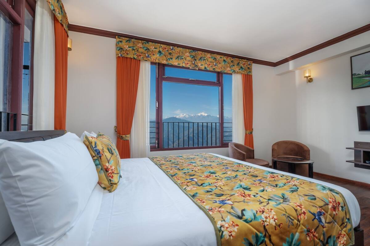 Quarto de hotel com cama, poltrona, mesinha, televisão e janela com vista para as montanhas.