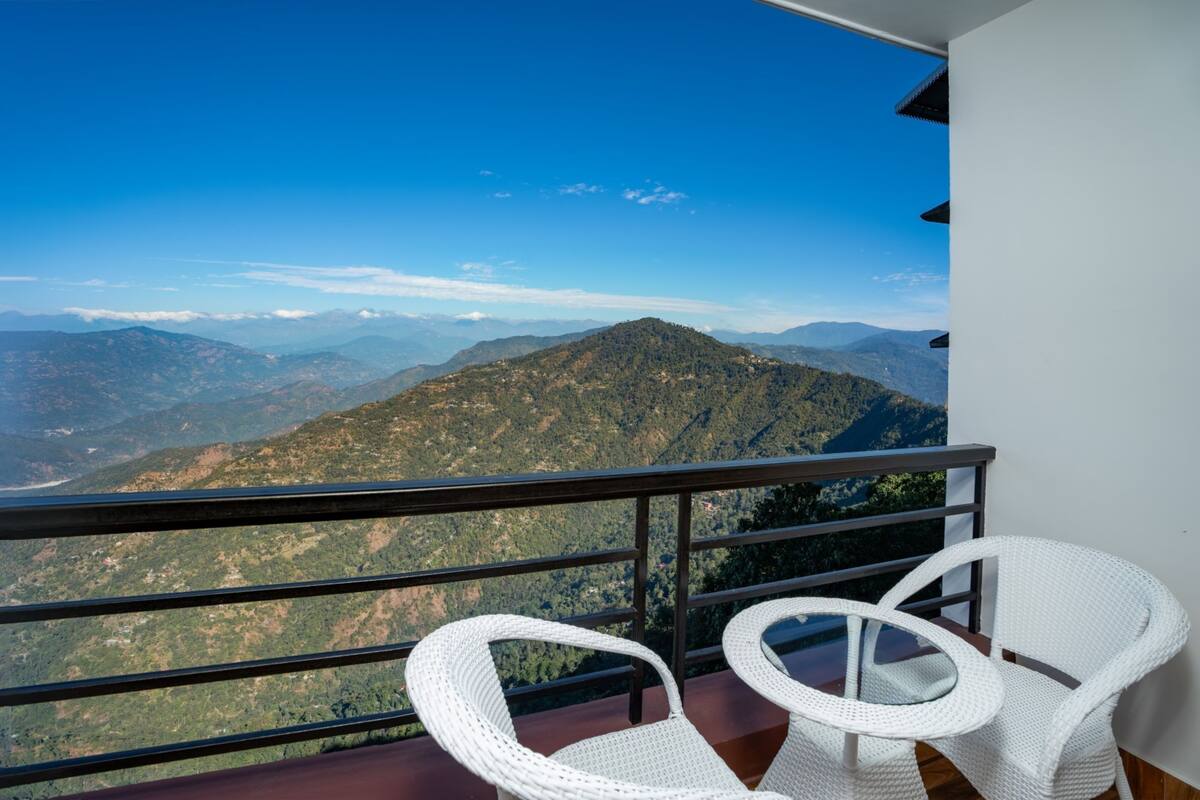 Varanda com duas cadeiras de vime brancas e uma mesa de vidro, com vista para uma paisagem montanhosa.