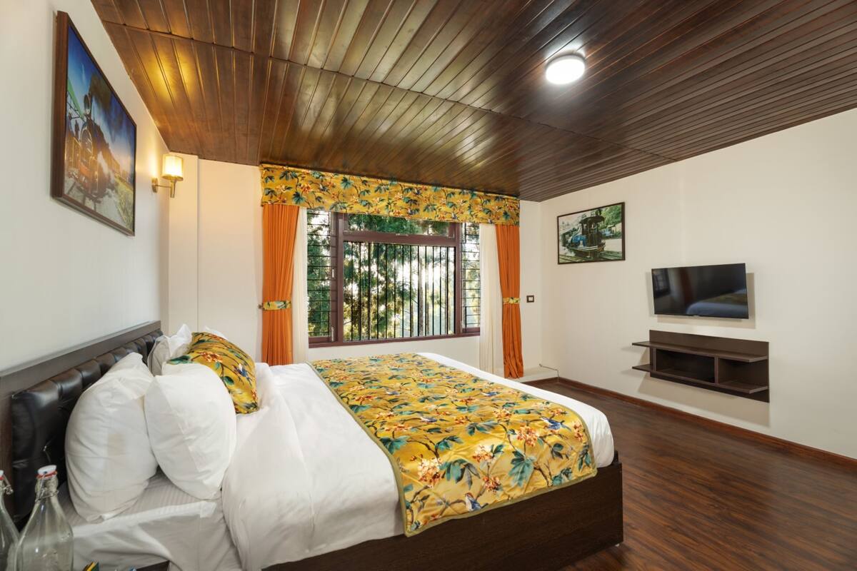 Quarto com teto de madeira, cama com colcha florida, televisão na parede e janela com cortinas.