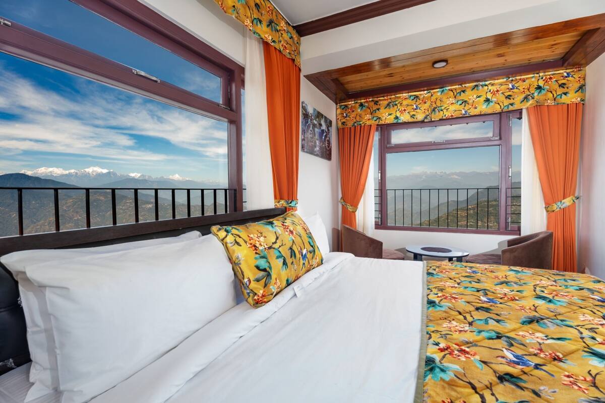 Quarto de hotel com uma cama, uma pequena mesa e vista para montanhas através de grandes janelas.