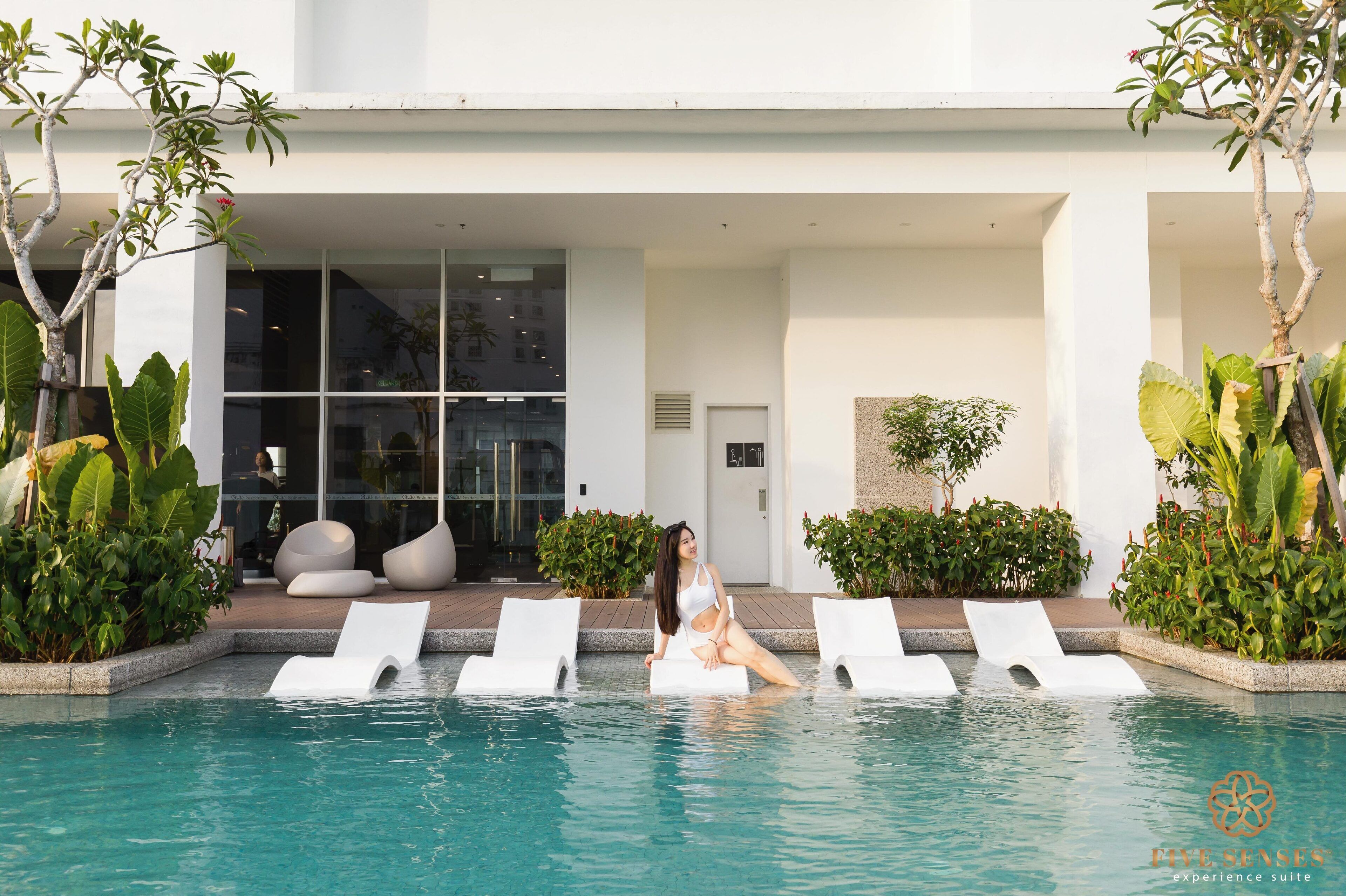 Área de piscina com espreguiçadeiras e uma pessoa sentada  à beira da piscina.