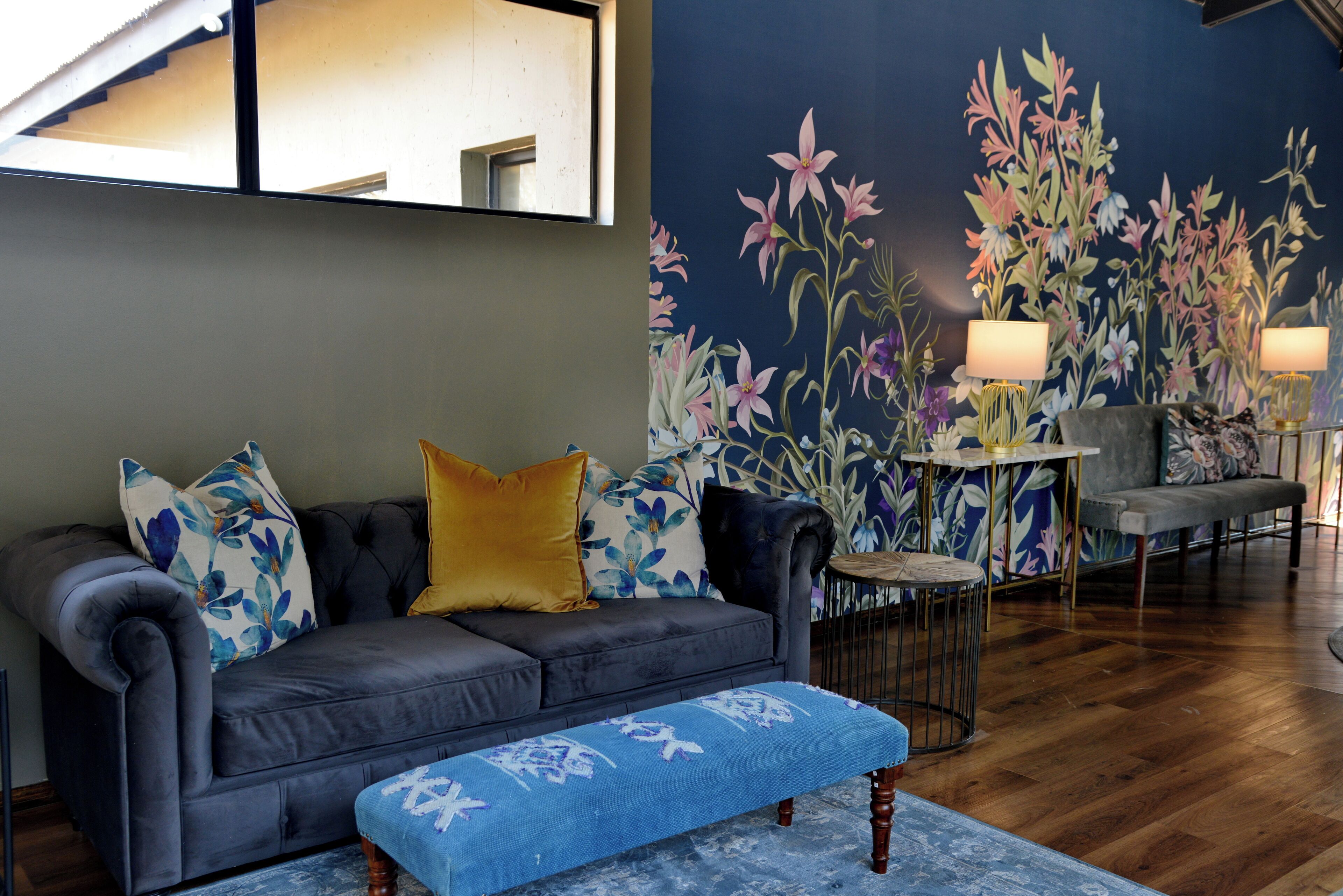 Uma sala de estar com um sofá cinzento, um banquinho azul estampado e um mural floral na parede.