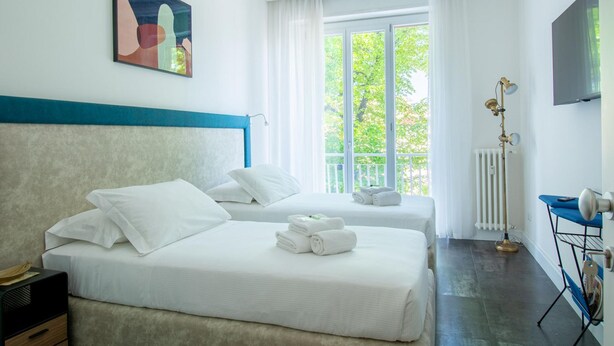 Quarto de hotel com duas camas, um quadro na parede, uma janela grande com cortinas, um radiador e uma TV fixada na parede.