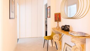 Um corredor com uma porta branca, um espelho com moldura dourada, uma mesa com objetos decorativos e uma cadeira amarela.