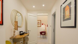 Um corredor com espelho, uma cadeira vermelha e porta que dá para outro quarto.