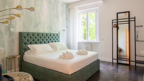 Cama bem arrumada com lençóis e toalhas brancos, cabeceira estofada verde e uma janela grande com vista para o verde lá fora.