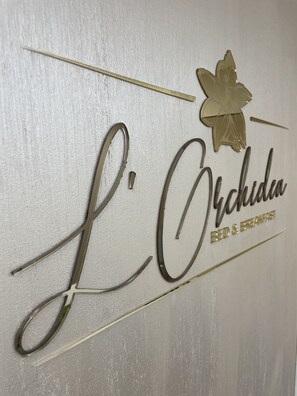 Placa na parede com o nome 'L'Orchida Bed & Breakfast' em fonte cursiva, acompanhada de uma flor dourada decorativa.