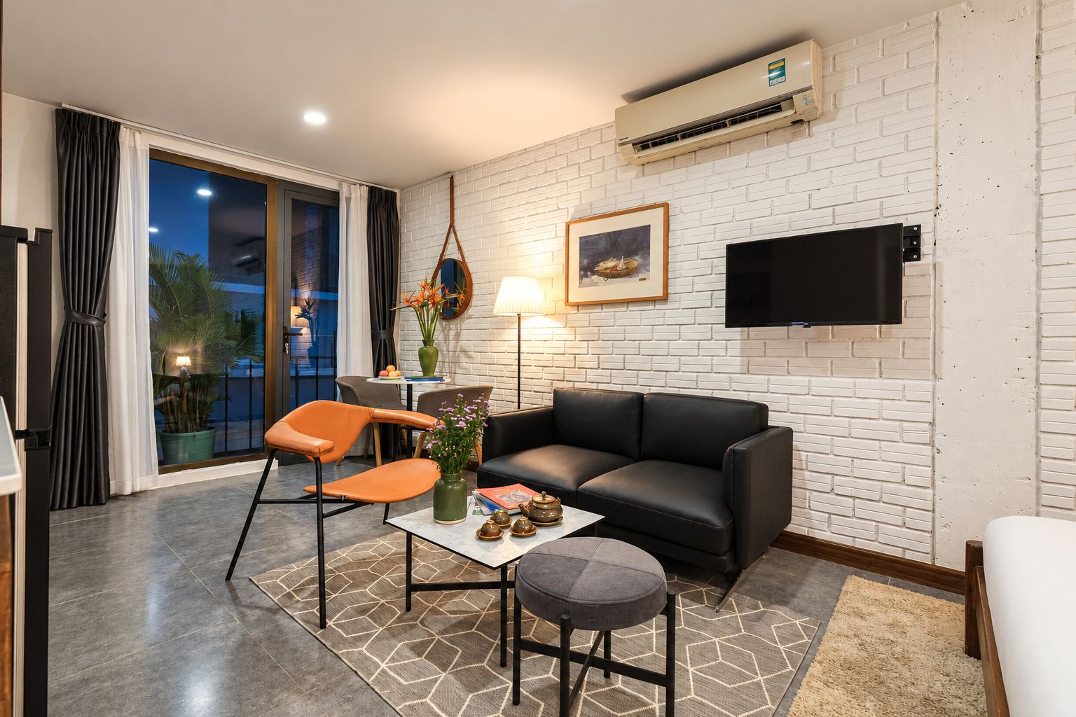 Uma sala de estar moderna com um sofá de couro preto, uma mesa de centro redonda e uma poltrona laranja. Há uma TV de tela plana na parede e um vaso de planta na mesa.